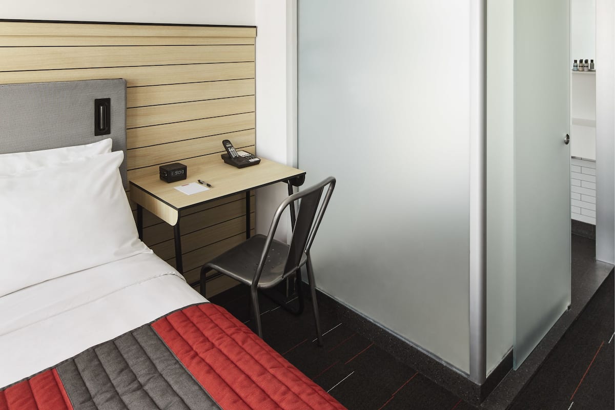 Quarto de hotel com cama, escrivaninha, cadeira, guarda-roupa e banheiro visível pela porta.