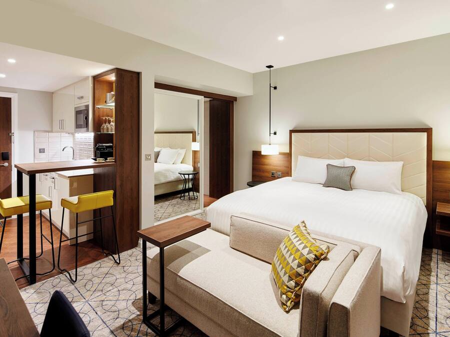 Quarto de hotel com cama, sofá, mesa pequena, área de cozinha e banheiro visível por uma porta aberta.