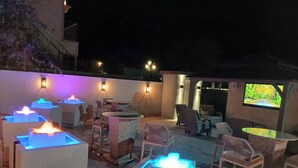 Uma área de estar ao ar livre com mesas e cadeiras iluminadas, uma tela de televisão exibindo uma paisagem natural e uma escadaria que dá acesso a um andar superior.