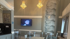 Uma ampla área de refeições com piso de mármore, cadeiras modernas e mesas. Há uma televisão montada na parede e um painel decorativo.