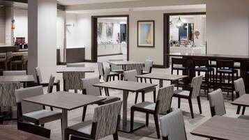 Uma área de refeições moderna com várias mesas e cadeiras, um bar e vista para a cozinha ao fundo.