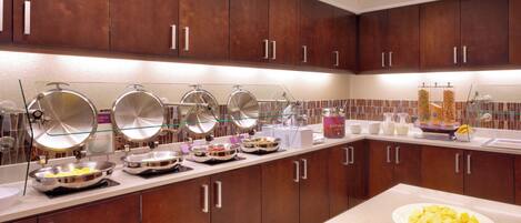 Uma mesa de buffet organizada com várias estações de serviço, incluindo recipientes com comida e diversos utensílios.