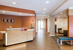 Uma área de recepção de hotel com um balcão de madeira, um letreiro com a palavra 'Hello', um mapa na parede e uma área de estar com um banco.