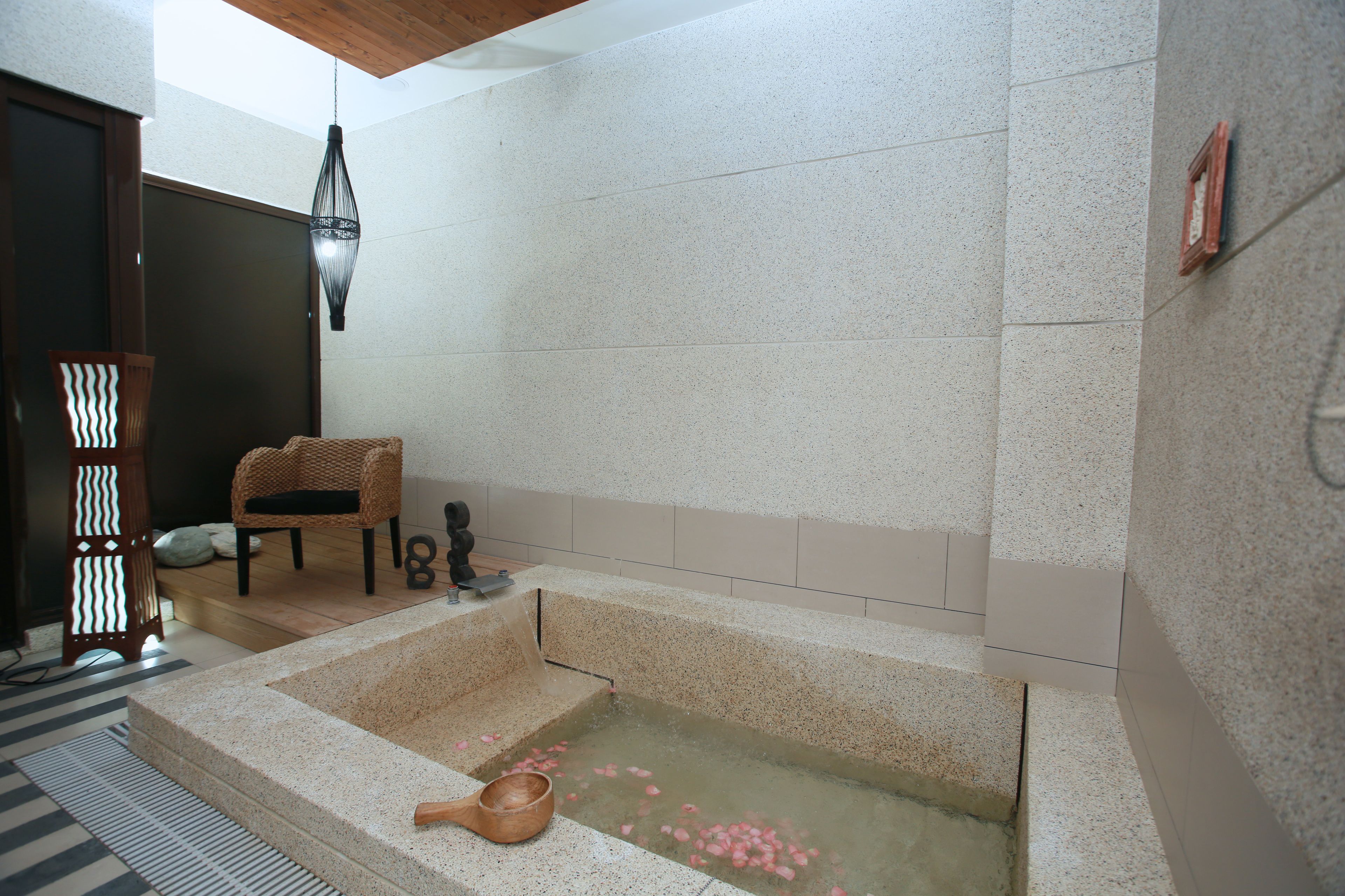 Uma sala de spa com uma banheira de pedra, uma bacia de madeira e uma cadeira de fibra natural.