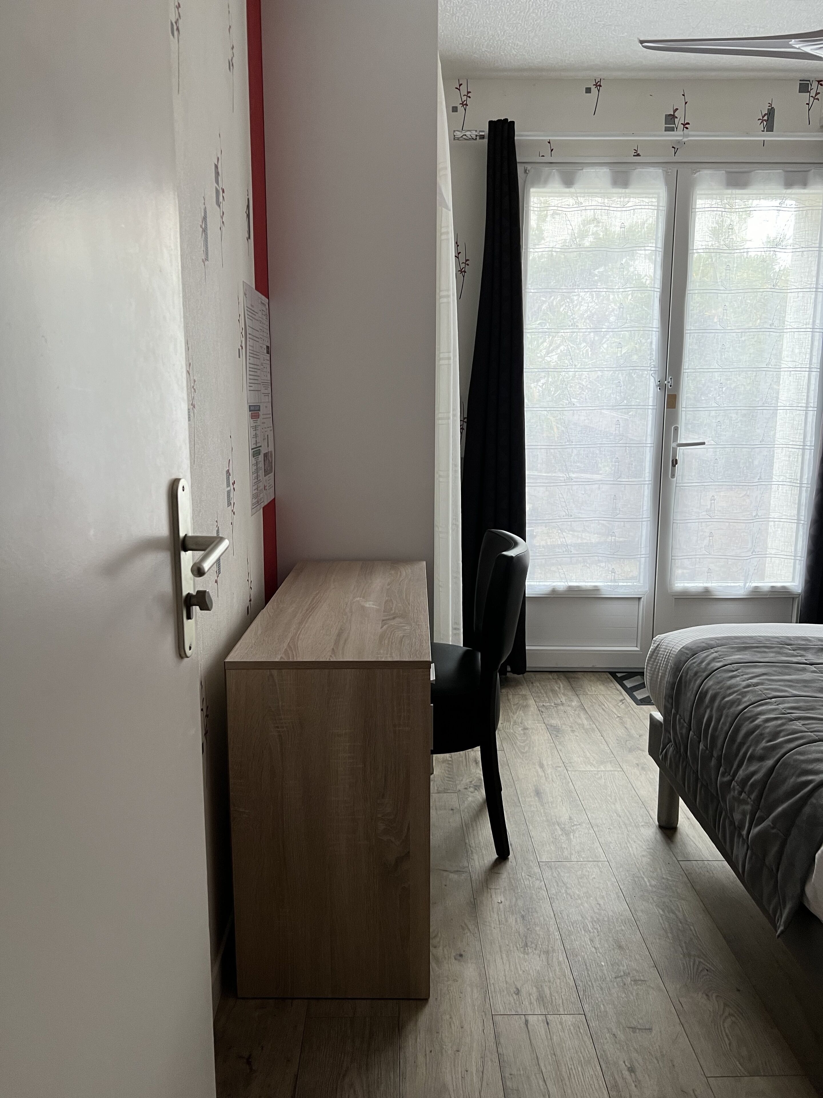 Um quarto com escrivaninha de madeira, uma cadeira preta, uma cama e uma janela com cortinas.