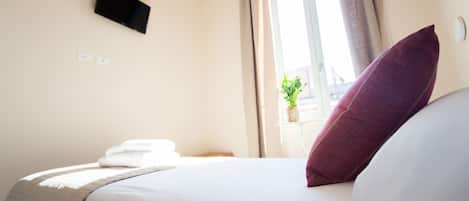 Cama bem arrumada com edredom branco e uma almofada vermelha, televisão de tela plana na parede e janela com cortinas.