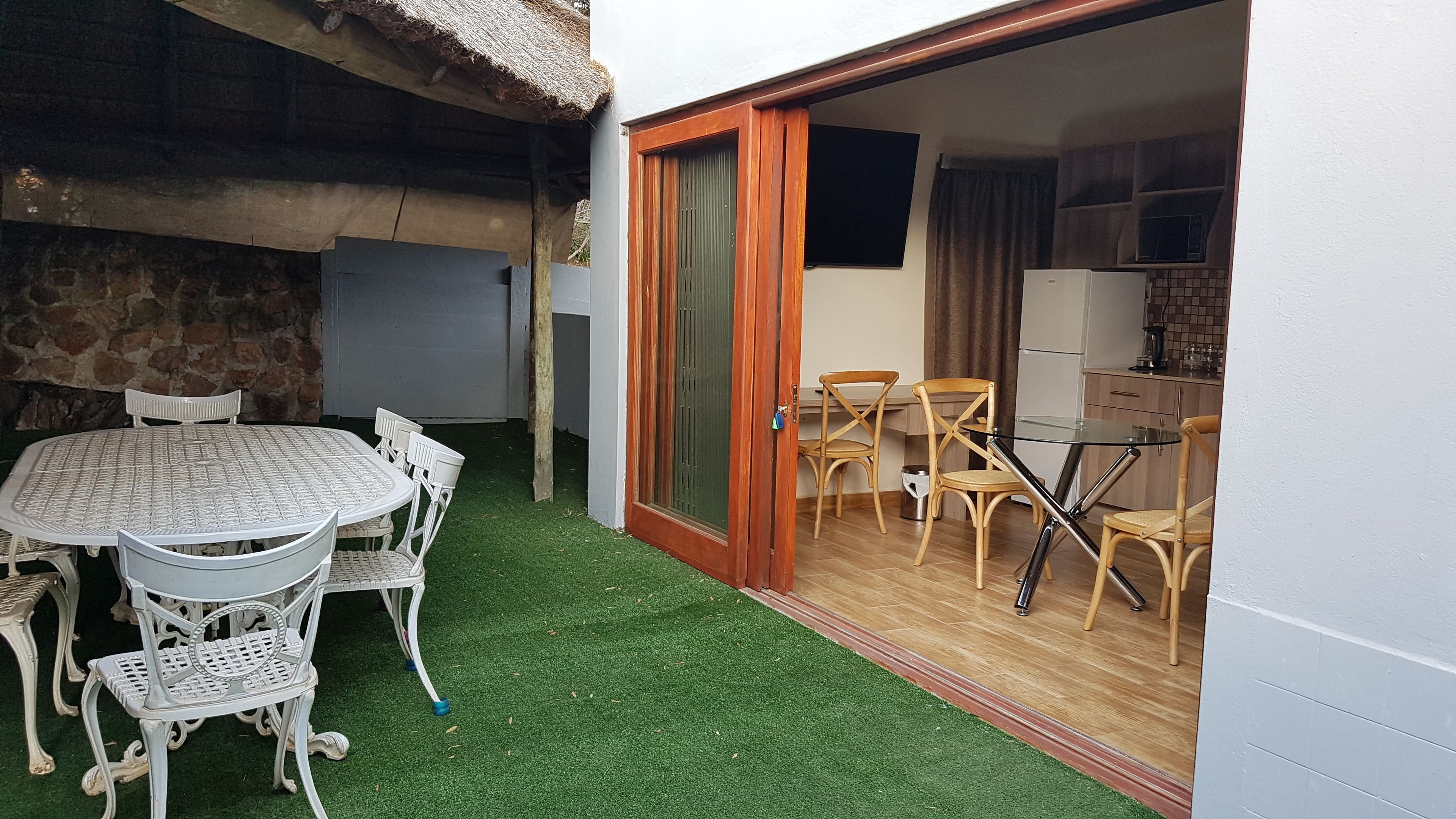 Um pátio com mesa redonda e cadeiras brancas, porta de madeira que dá para uma cozinha e parede de pedra.