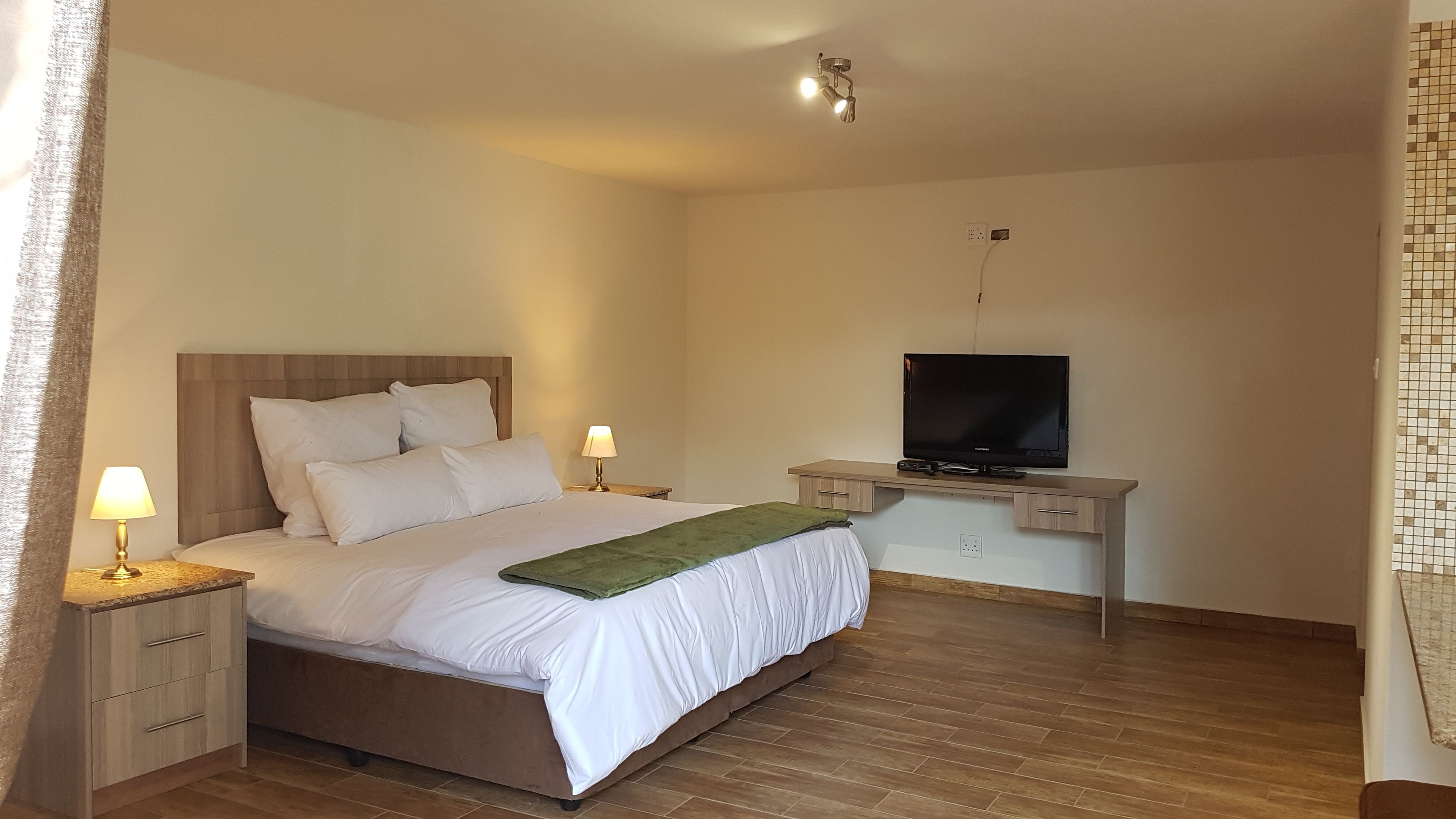 Quarto de hotel bem arrumado com cama, abajures, TV em suporte de madeira e manta verde.