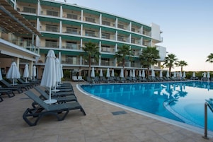 Uma área de piscina com espreguiçadeiras e guarda-sóis, um edifício com várias andares e varandas, e palmeiras.