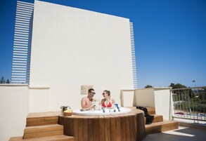 Um casal relaxando em uma banheira de hidromassagem numa varanda, com um céu azul claro.