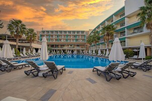 Área de piscina de hotel com espreguiçadeiras e guarda-sóis, cercada por palmeiras e um edifício com várias varandas.