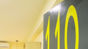 Quarto de hotel com porta identificada com o número 110, uma pequena mesa com uma toalha, um cesto de lixo, um lustre pendurado e um ar condicionado fixado na parede.
