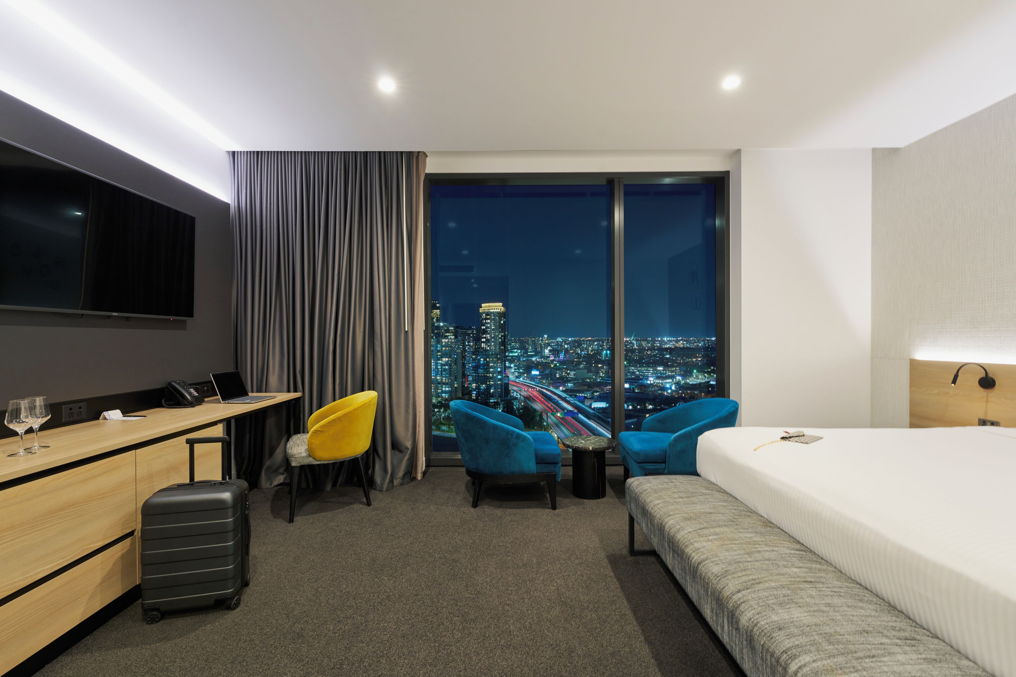 Quarto de hotel com cama, escrivaninha, cadeiras e vista noturna da cidade.