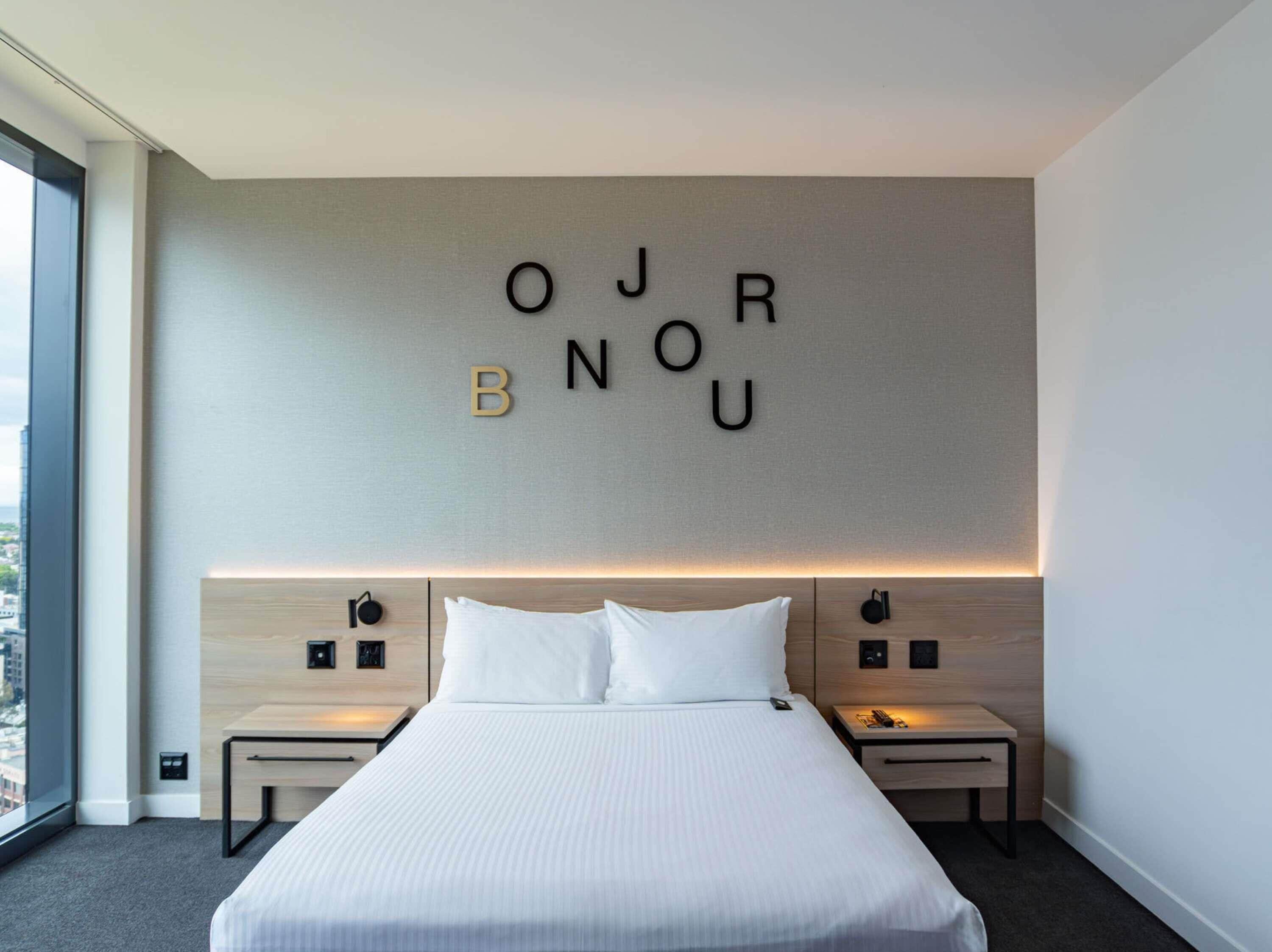 Quarto de hotel moderno com uma cama grande, duas mesas de cabeceira e um letreiro fixado na parede com a inscrição "OJR B NOU".