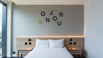 Quarto de hotel moderno com uma cama grande, duas mesas de cabeceira e um letreiro fixado na parede com a inscrição "OJR B NOU".