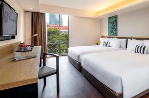 Quarto de hotel com escrivaninha, duas camas, televisão e uma janela grande com vista para edifícios e árvores.