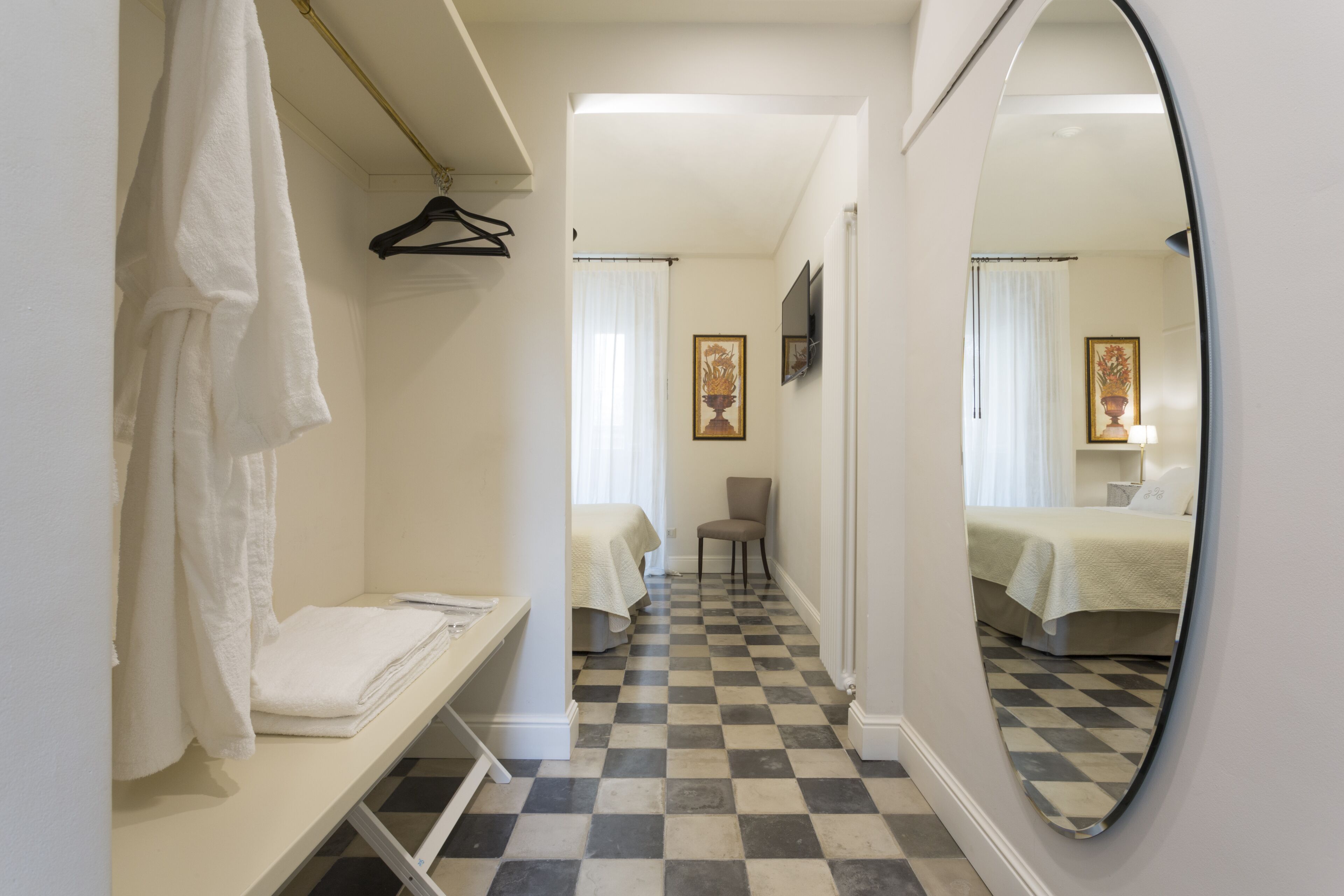 Quarto de hotel com chão xadrez, cama com colcha branca, uma cadeira, espelho e porta-toalhas.