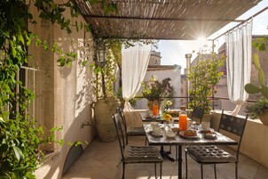 Pátio ensolarado com mesa posta para o café da manhã, cercado por plantas em vasos e uma pérgula.