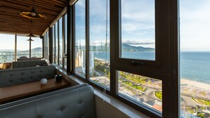 Interior moderno com grandes janelas, teto de madeira e área de estar com vista para o mar e a cidade.