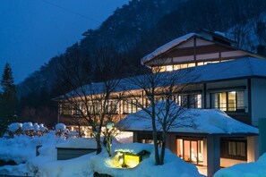 Um edifício moderno com várias janelas e telhado inclinado, rodeado por árvores cobertas de neve e montanhas ao fundo.
