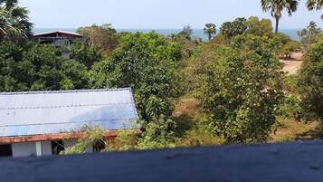 Um edifício com telhado de metal corrugado, rodeado por vegetação densa e palmeiras, com vista clara para o oceano ao fundo.