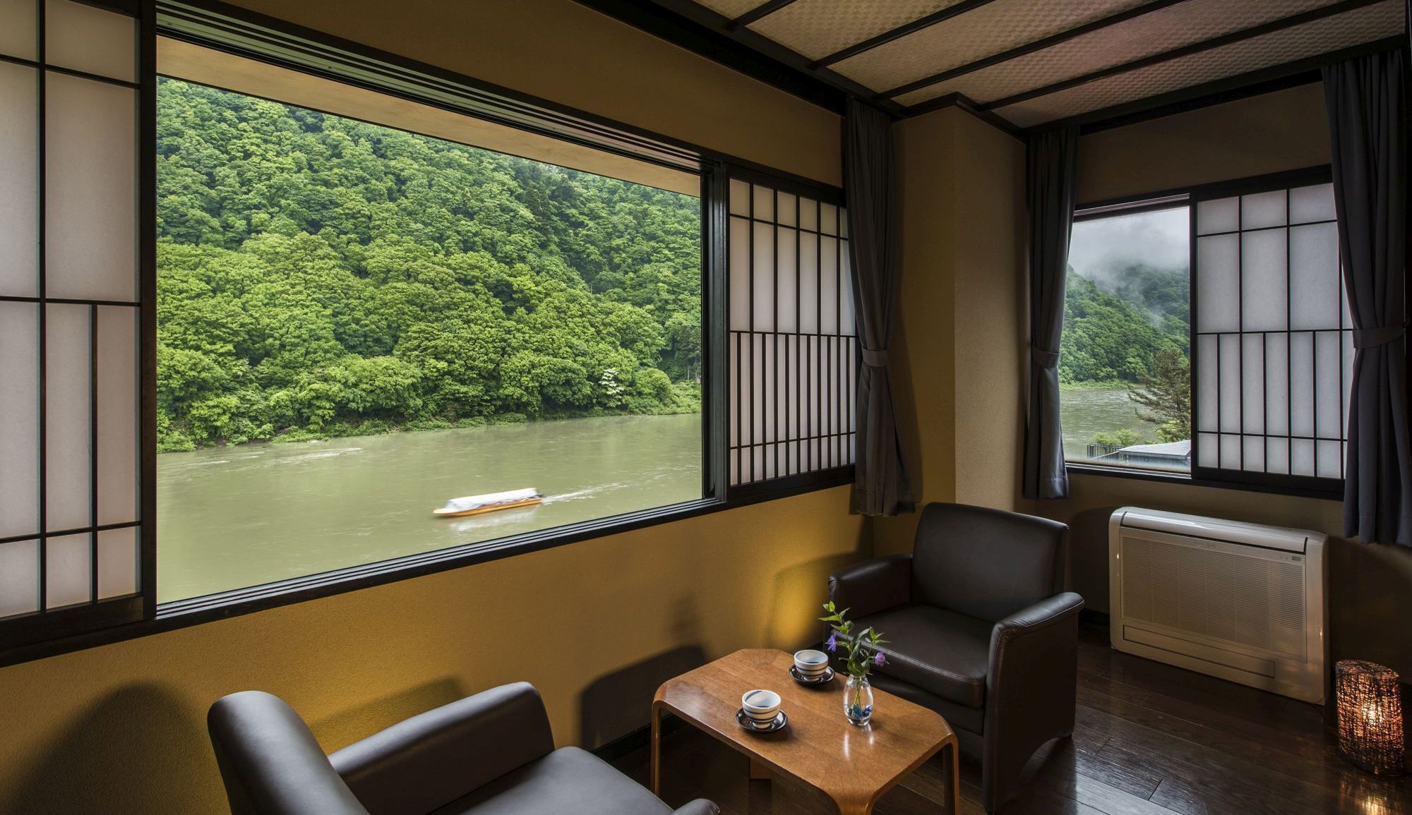 Quarto com piso de tatame, janela tradicional japonesa com portas shoji, vista para um rio e uma mesa com um vaso e tigelas.