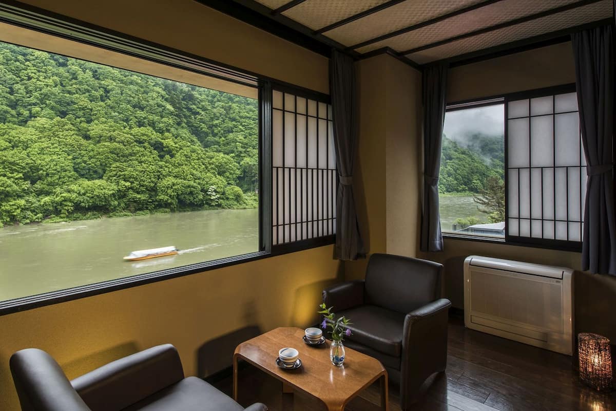 Quarto com piso de tatame, janela tradicional japonesa com portas shoji, vista para um rio e uma mesa com um vaso e tigelas.