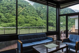 Quarto com uma janela grande de frente para um rio e colinas arborizadas. Há um sofá azul e uma mesa de madeira com um tapete.