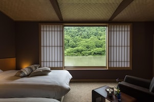 Quarto de hotel com uma grande janela, uma cama, uma cadeira e uma mesa pequena com um bule e xícara.
