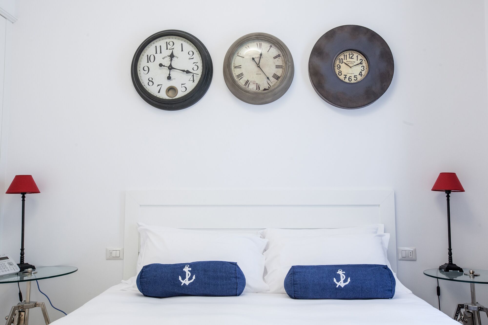 Quarto de hotel com uma cama, duas luminárias vermelhas e três relógios de parede.