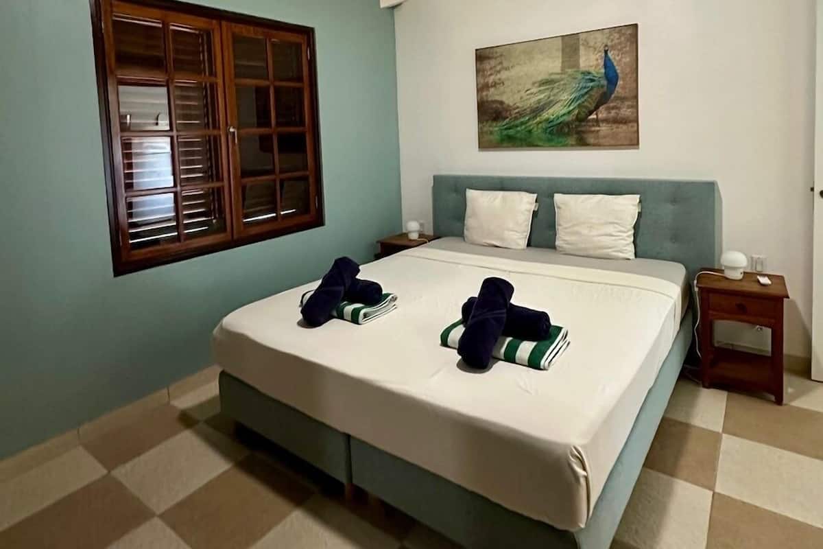 Quarto com cama, ventoinha no teto, janela com persianas, um quadro e mesa de cabeceira.