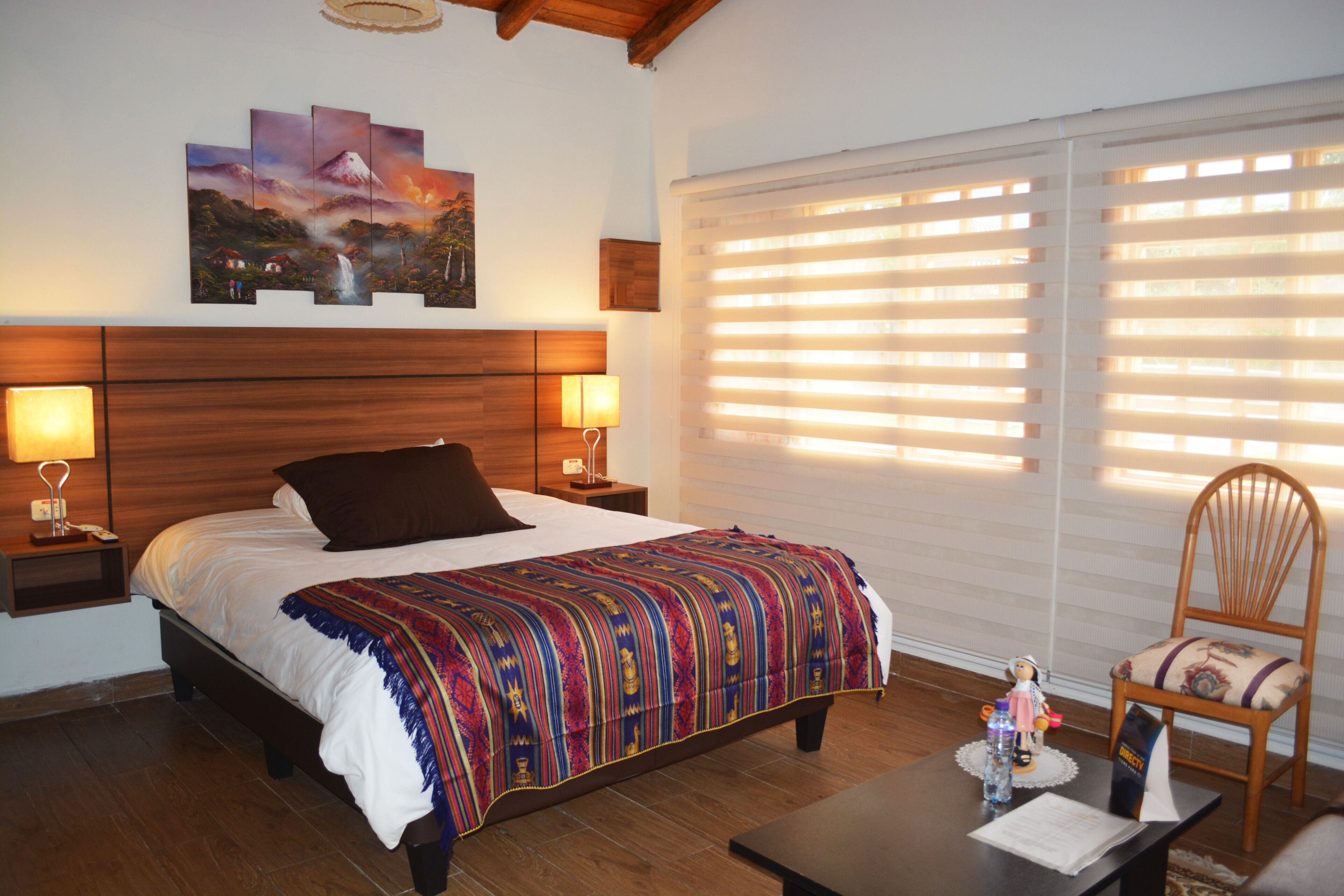 Quarto de hotel bem arrumado com uma cama grande, cabeceira de madeira, duas luminárias de cabeceira, uma mesa de cabeceira de madeira, uma pequena mesa com uma garrafa e uma estatueta, e uma janela com persianas.