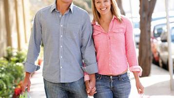 Um casal caminhando pela calçada, de mãos dadas e carregando sacolas de compras.