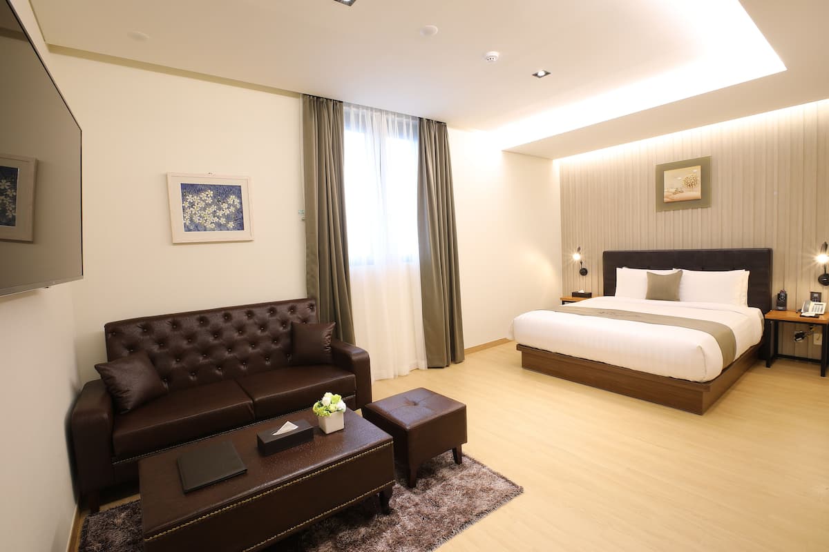 Quarto de hotel moderno com sofá de couro marrom, mesa de cabeceira de madeira, cama grande com roupa de cama branca e pequena mesa lateral com abajur.