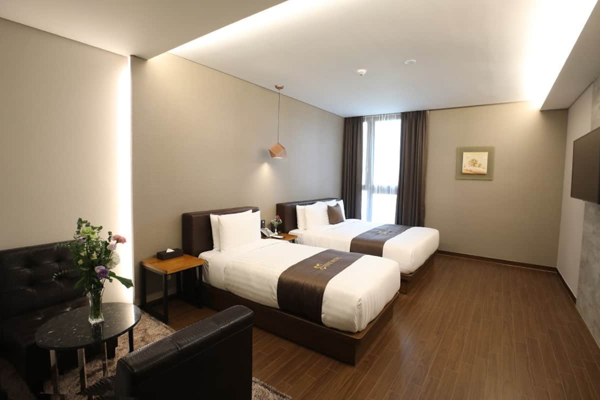 Quarto de hotel com duas camas, um sofá, uma mesinha com um vaso de flores, uma televisão e uma janela com cortinas.