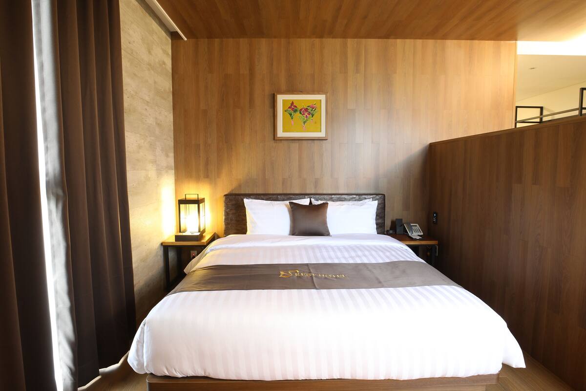 Quarto de hotel com uma cama grande, cabeceira de madeira, mesa de cabeceira com abajur e um quadro na parede.