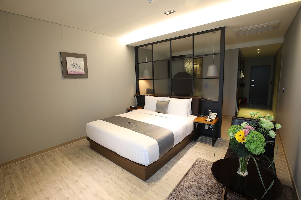 Quarto de hotel moderno com uma cama grande, mesa de cabeceira com abajur, uma pequena mesa com um vaso de flores e um quadro na parede.