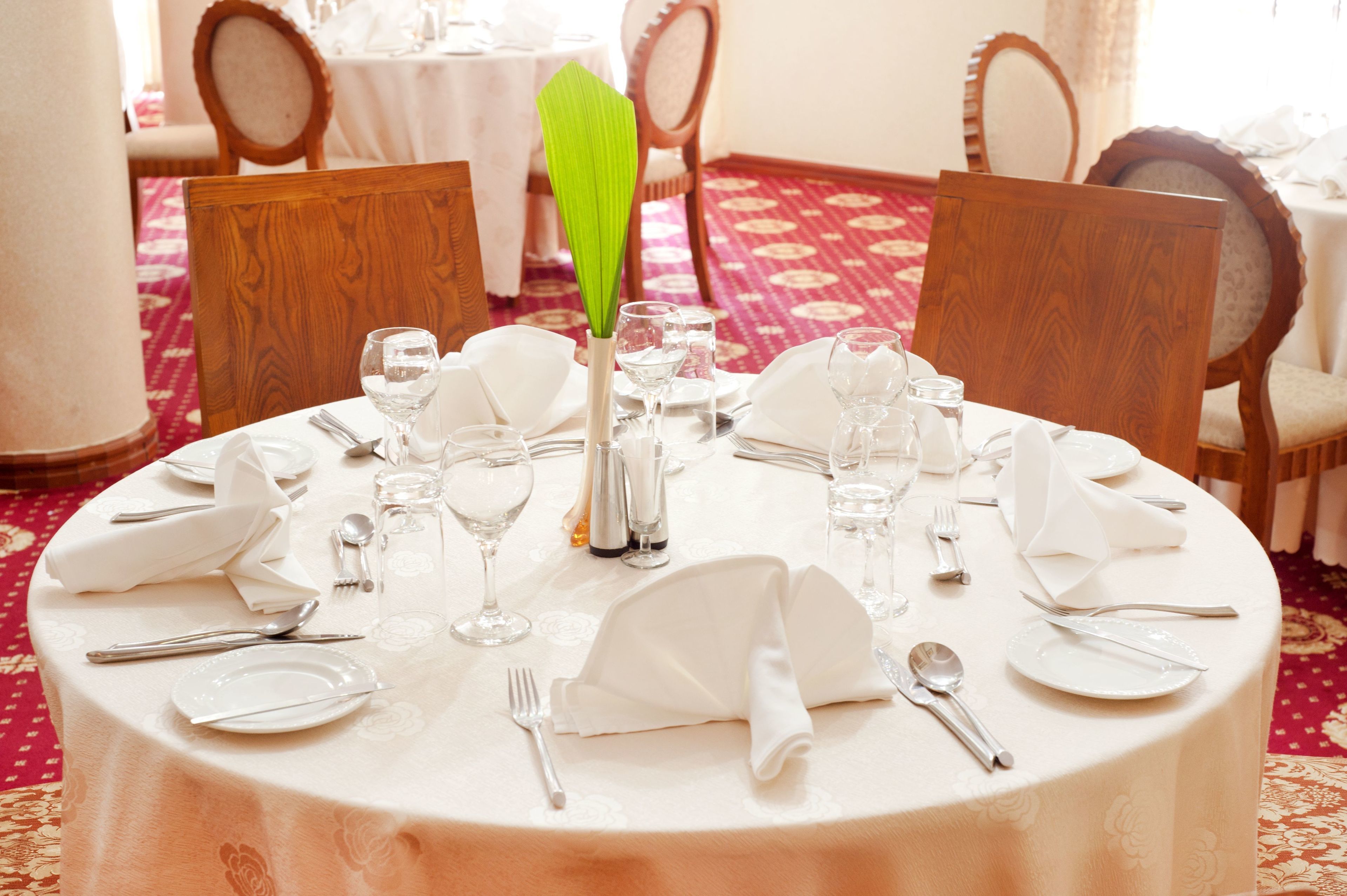 Mesa de jantar redonda posta com louça branca, incluindo pratos, talheres e copos, em um ambiente com carpete estampado e cadeiras de madeira.