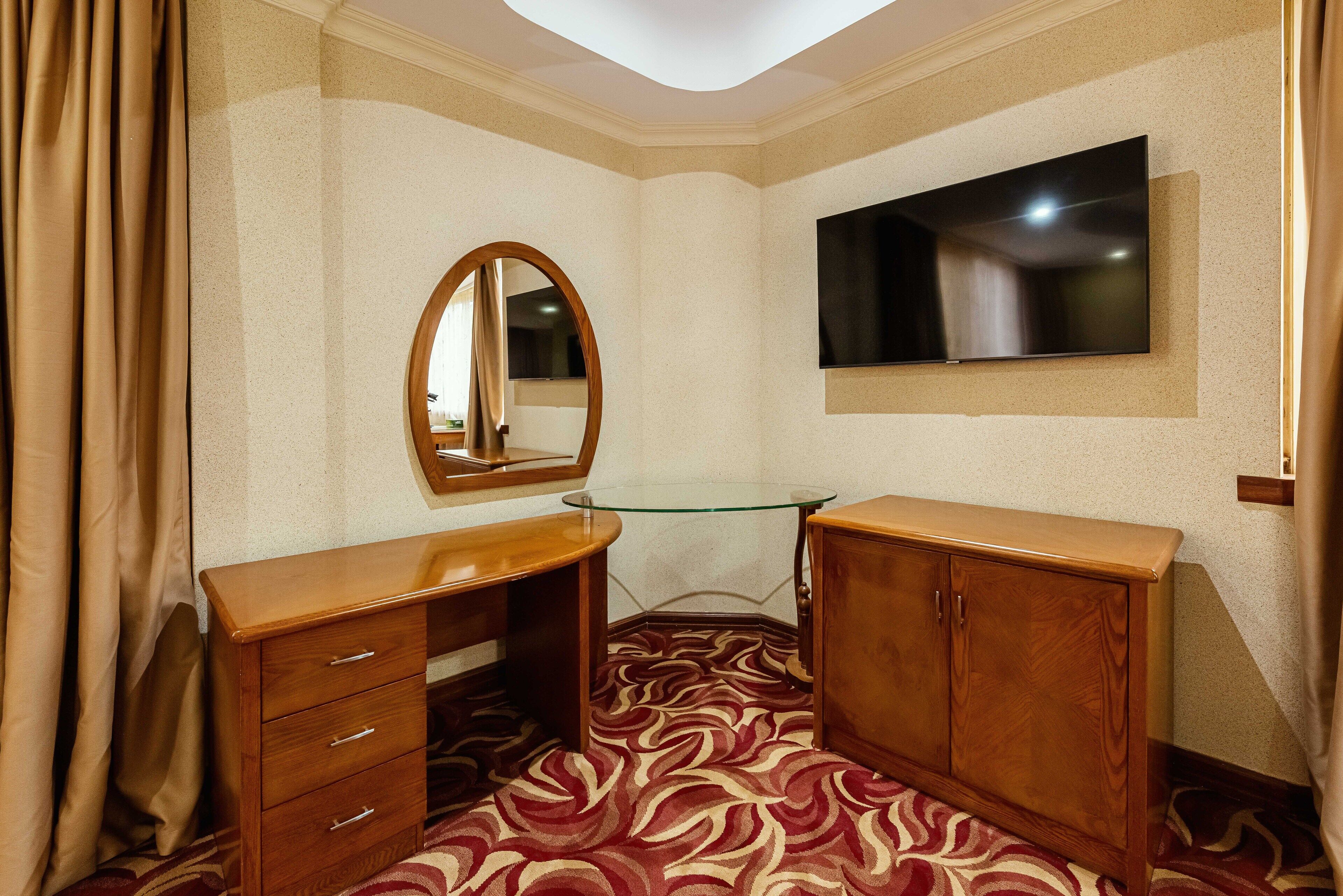 Quarto de hotel com escrivaninha de madeira, mesa com tampo de vidro, espelho, TV fixada na parede e carpete estampado.