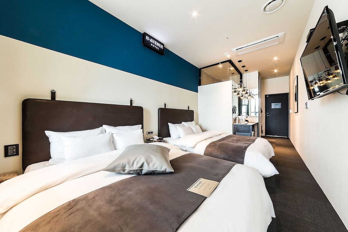 Quarto de hotel com duas camas, uma televisão de tela plana e uma placa indicando "Havenue Hotel".