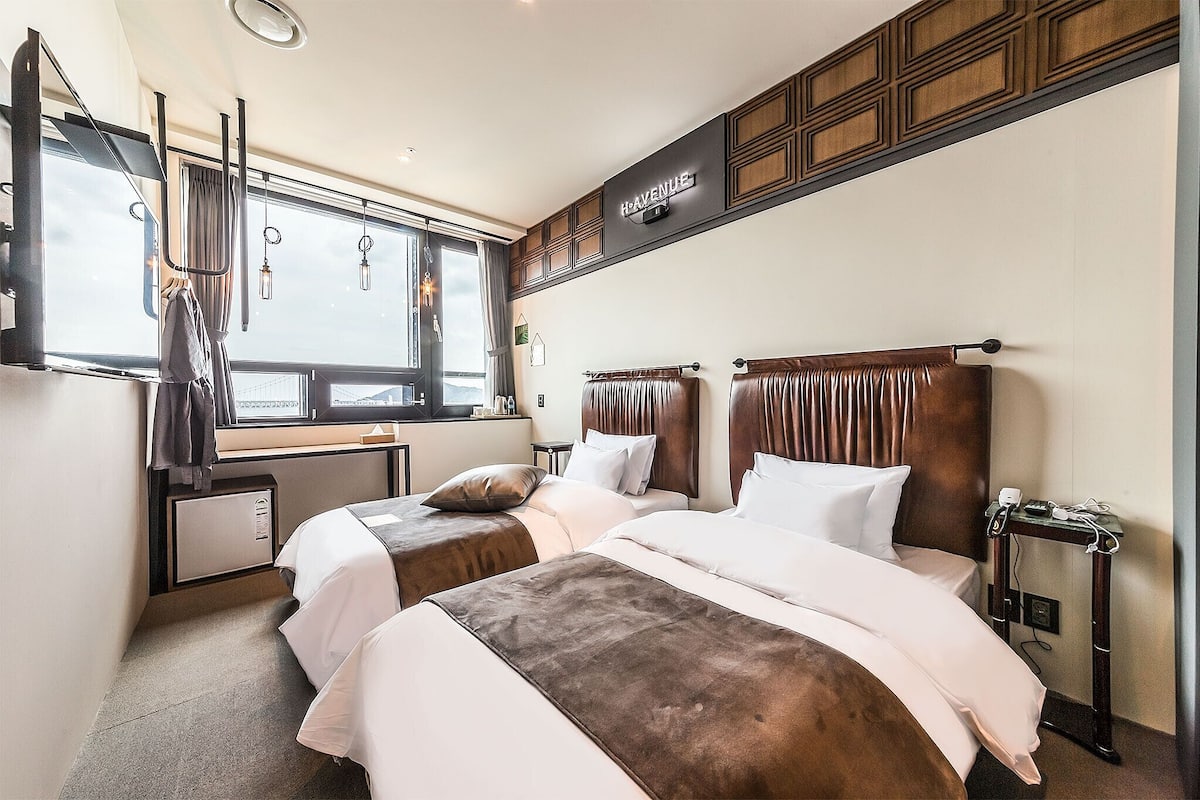 Quarto de hotel com duas camas, uma escrivaninha pequena, uma janela com cortinas e uma placa na parede.