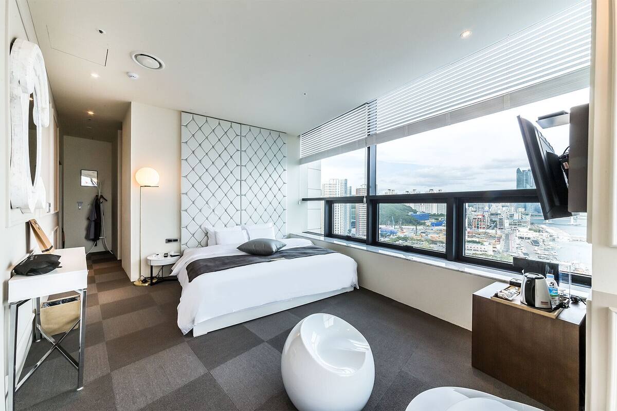 Quarto de hotel moderno com uma cama grande, uma escrivaninha, uma cadeira, um espaço de estar circular e vista para a cidade através de janelas amplas.