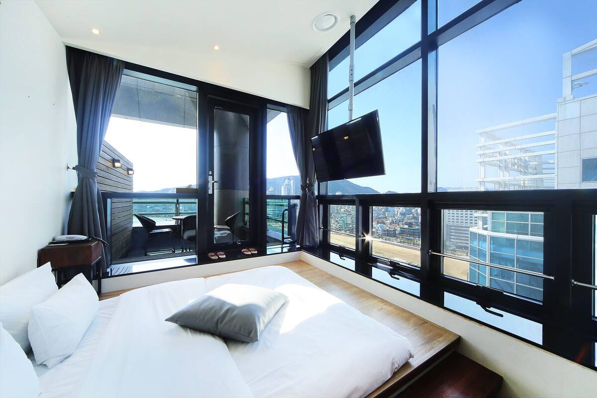 Quarto de hotel moderno com uma cama grande, uma TV e varanda com vista para a cidade.