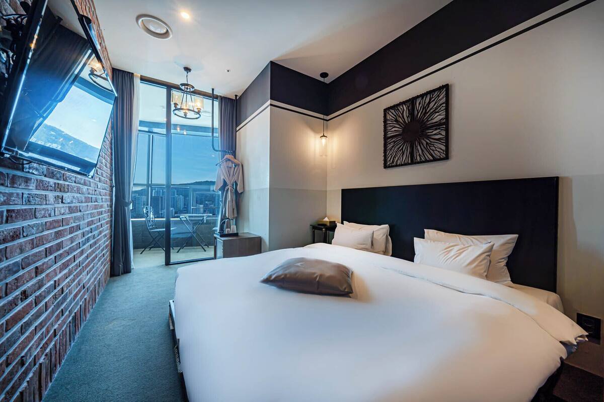 Quarto de hotel com uma cama grande, vista através de uma porta de vidro, parede de tijolos e luminária moderna.