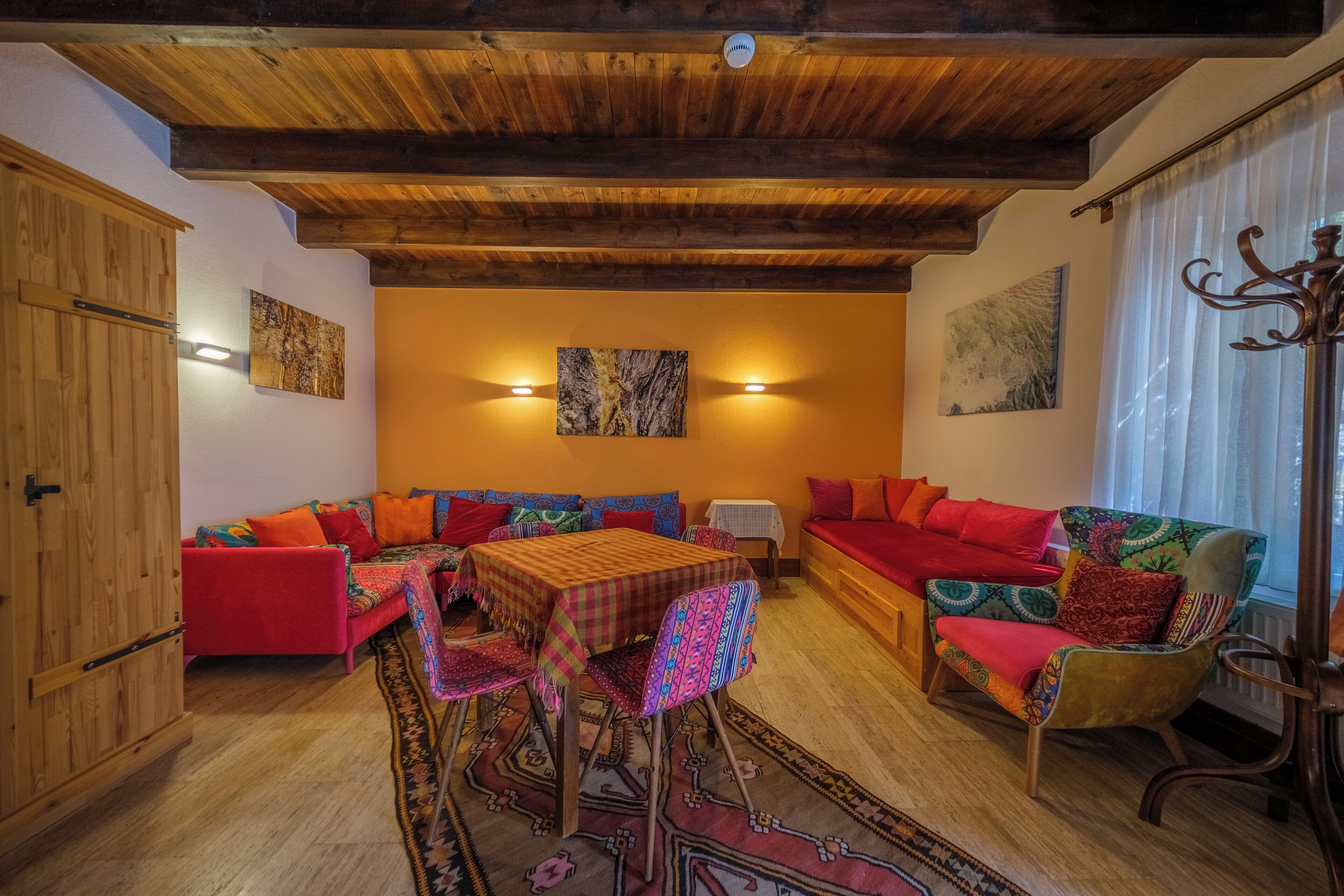 Uma sala de estar aconchegante com vigas de madeira no teto, um sofá vermelho, cadeiras coloridas e um tapete estampado.