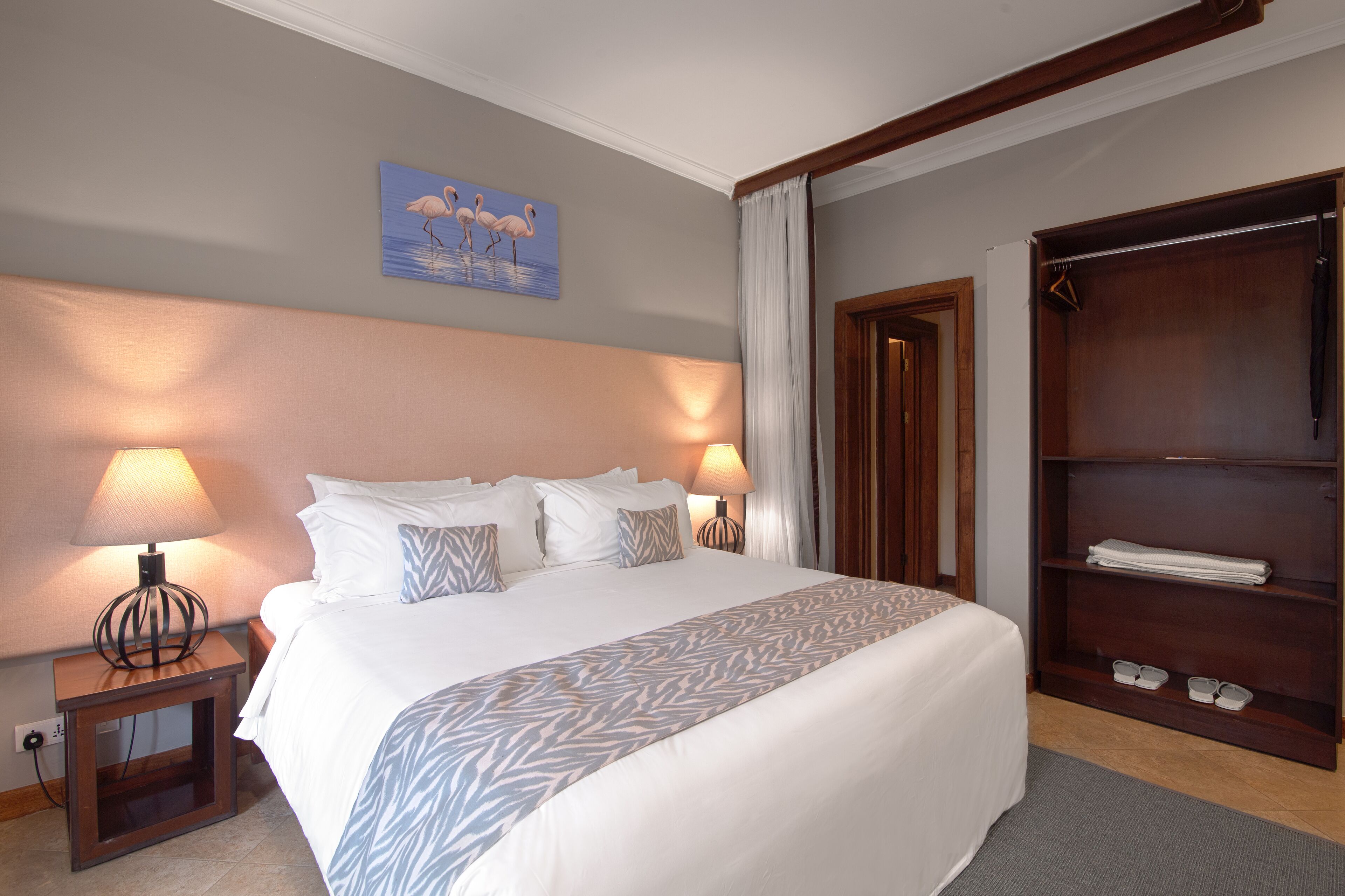 Quarto de hotel com uma cama grande, duas luminárias de cabeceira, um guarda-roupa de madeira e um quadro com flamingos na parede.