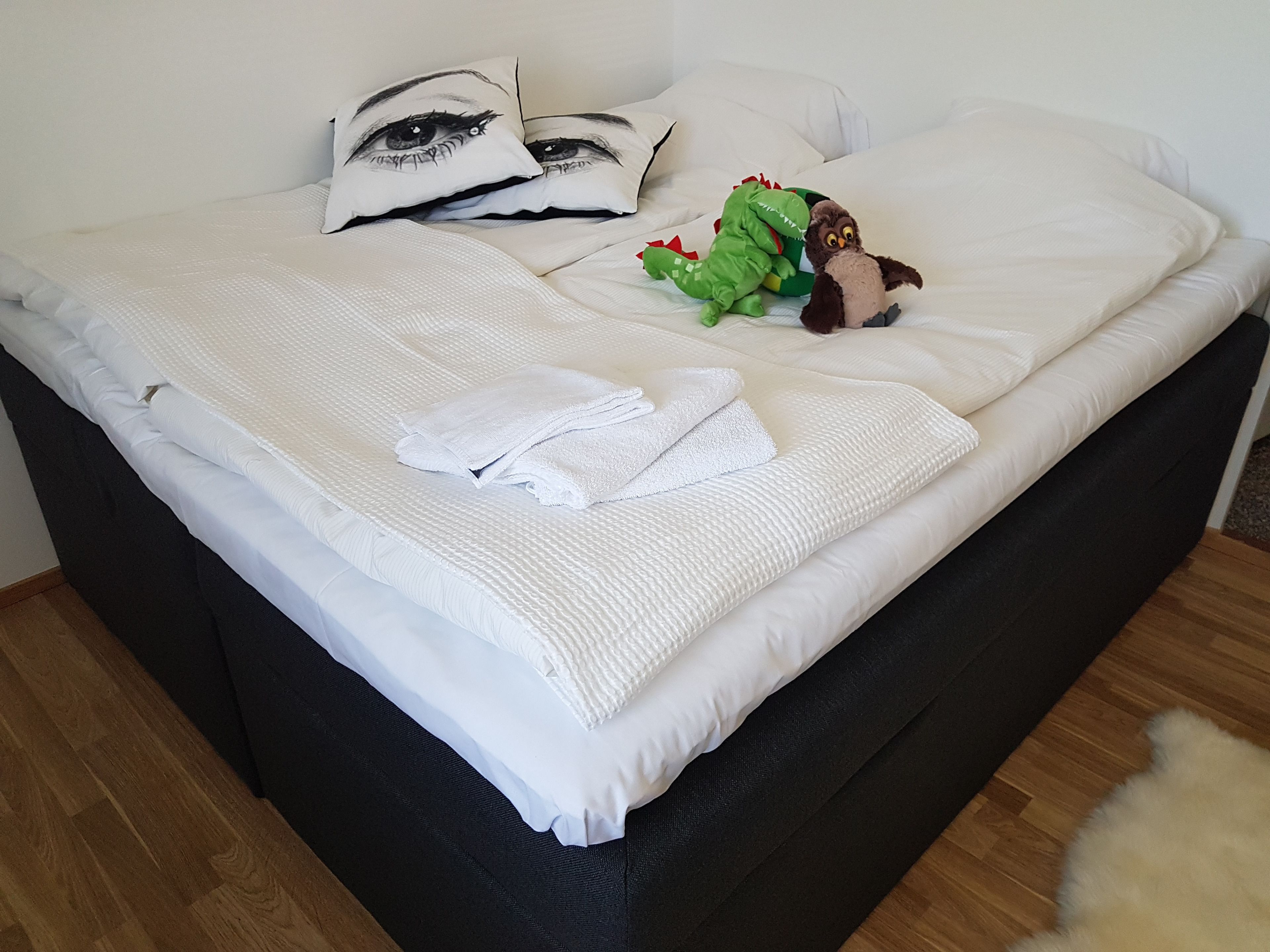 Cama bem arrumada com roupa de cama branca, duas almofadas com estampa de rosto, um brinquedo de pelúcia verde e um brinquedo de pelúcia marrom.