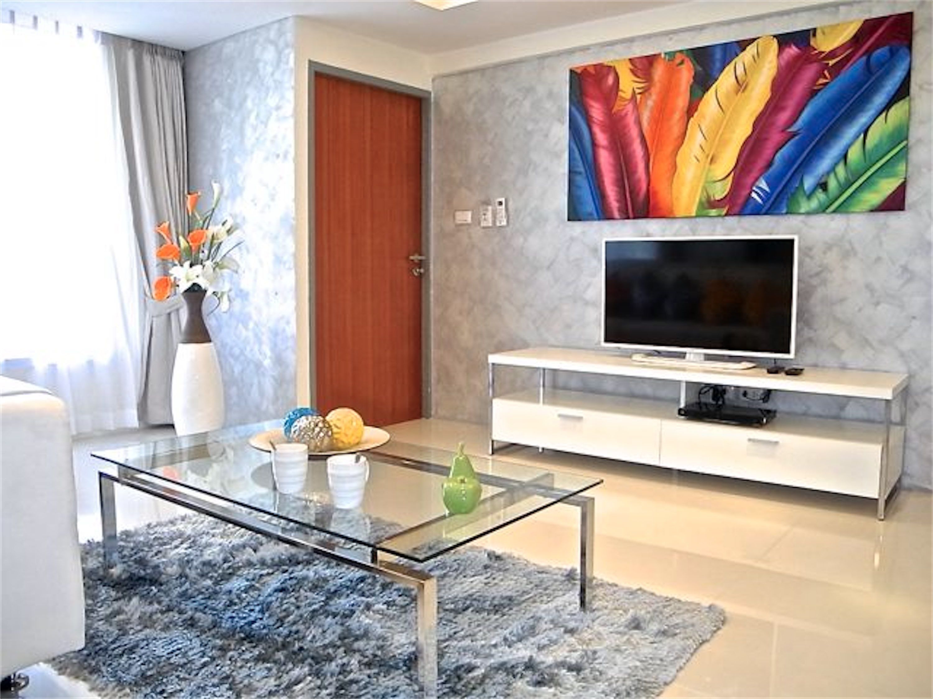 Uma sala de estar moderna com uma mesa de centro de vidro, um móvel branco para entretenimento com uma TV de tela plana, uma porta e uma pintura abstrata colorida na parede.