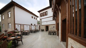 Um pátio externo com várias mesas e cadeiras, piso de pedra, e edifícios com fachadas e janelas de madeira.
