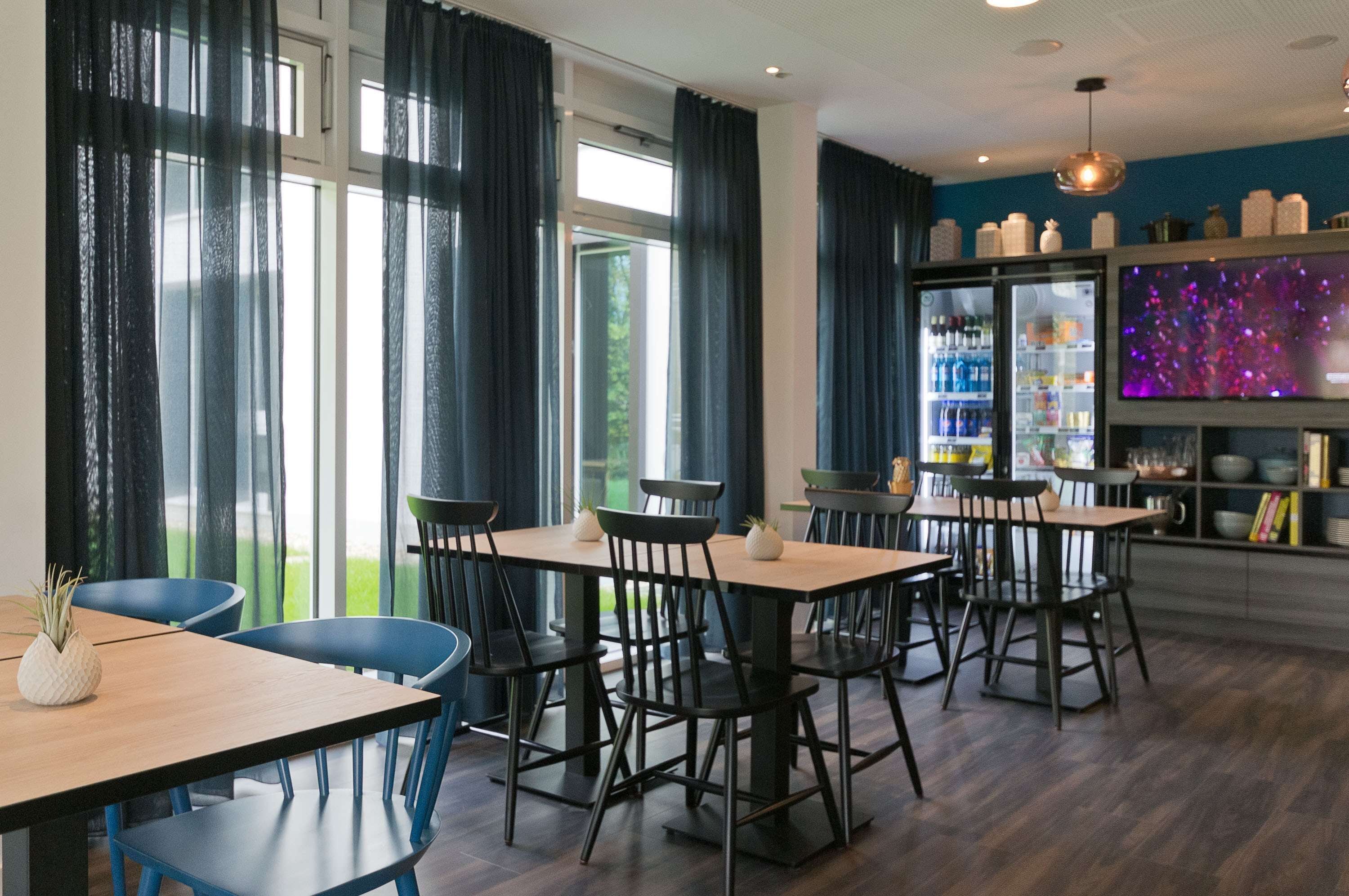 Um restaurante moderno com mesa de madeira e cadeiras azuis, janelas amplas com cortinas escuras e um espaço de bar com prateleiras de garrafas e copos.